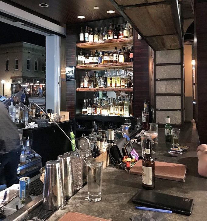 Bottles climb toward heaven on wooden shelves, while bartenders below craft liquid happiness for waiting patrons.