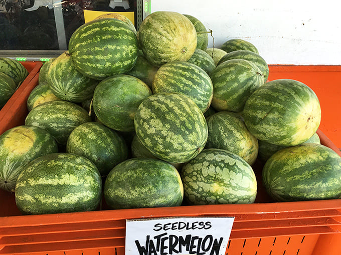 Seedless watermelons pile high in their orange crates, summer's juicy giants waiting to be chosen for picnics and poolside feasts.