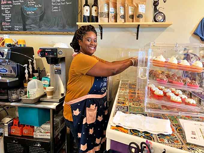 A staff member arranges freshly baked treats in the display case, her cat-adorned apron hinting at the establishment's unique mission.