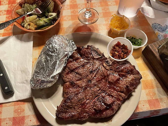 A perfectly mesquite-grilled steak dominates the plate, accompanied by a foil-wrapped baked potato that's practically begging to be unwrapped.