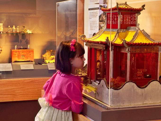 A young visitor discovers the magic of perspective, her wide-eyed wonder mirroring our own as she peers into an exquisite Chinese miniature palace.