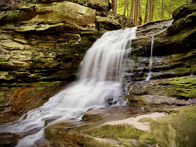 Water cascades down moss-covered rock faces, creating a refreshing mist that cools the air and adds magic to the forest atmosphere.