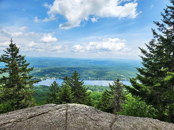 From the cliff's edge, the world opens to a sparkling lake, framed by trees and a perfect blue sky.