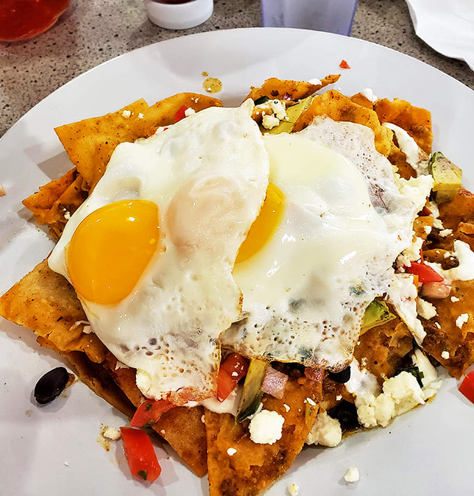 Chilaquiles Verde brings authentic Mexican breakfast vibes with crispy tortilla chips, perfectly fried eggs, and enough toppings to require architectural support.
