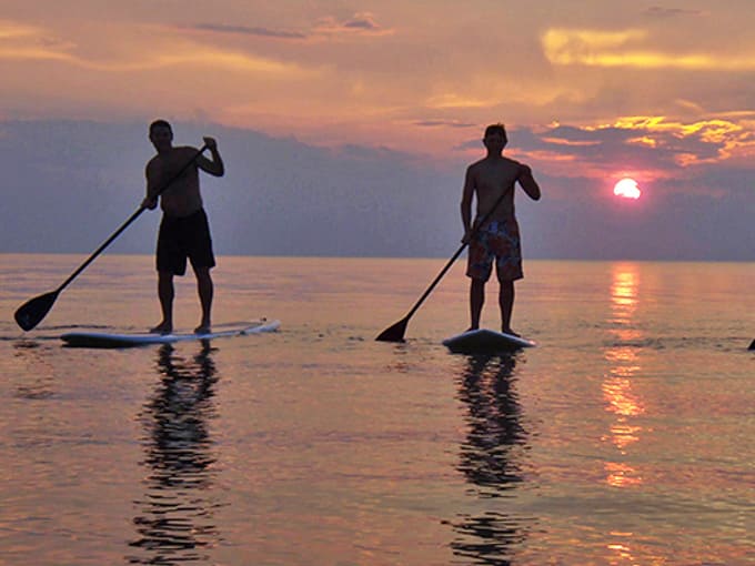Sunset paddleboarders glide across Lake Huron's mirror-like surface, where Michigan's sky puts on a spectacular color show to end the day.