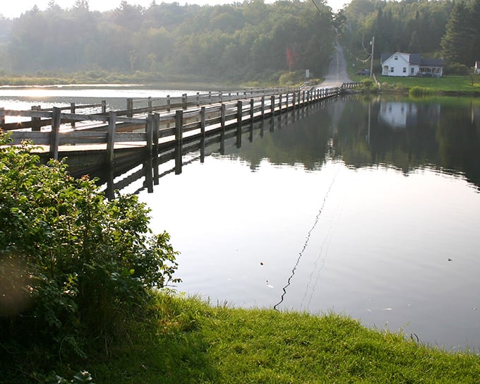 Morning mist hovers over Sunset Lake, where the wooden planks of Vermont's Floating Bridge create a pathway that seems to vanish into the fog.