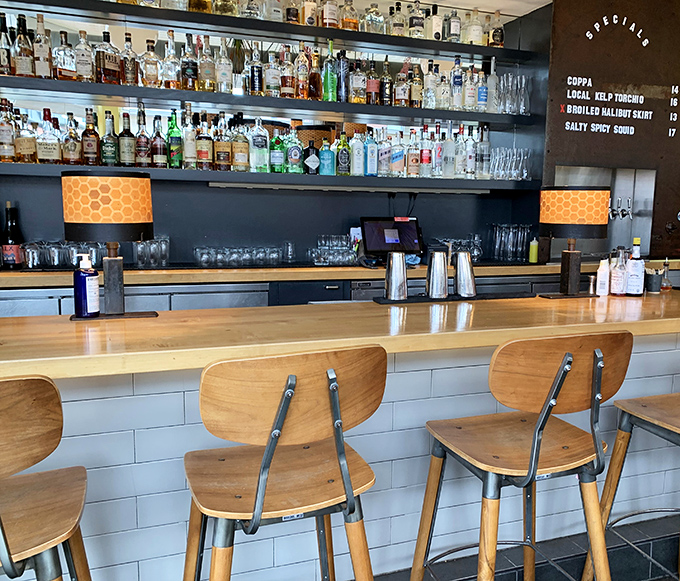 Bottles gleam like jewels against dark shelving while wooden barstools invite you to settle in for creative libations crafted with the same care as the food.