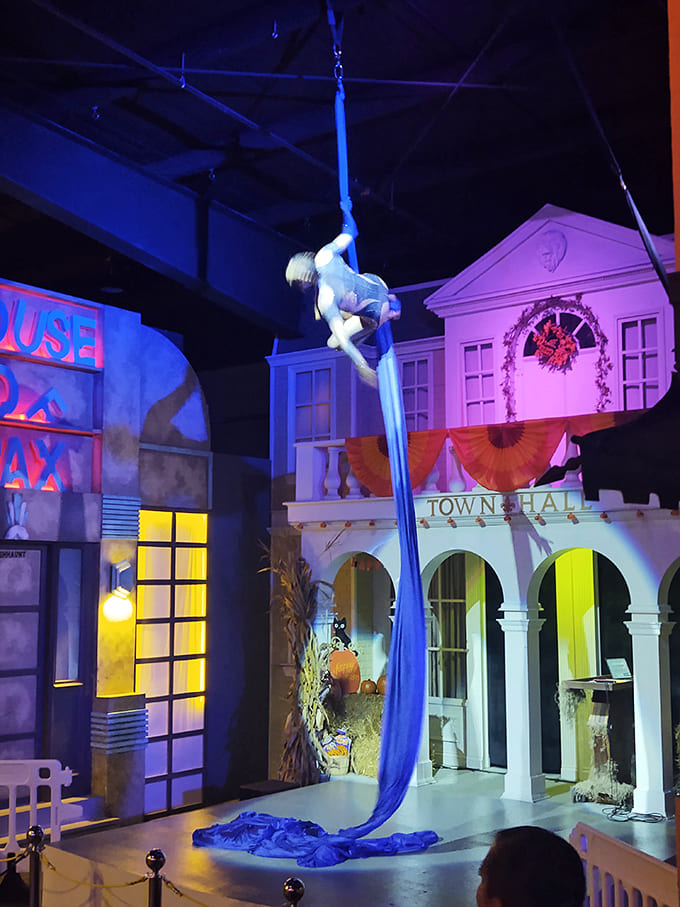 An aerial performer creates a moment of macabre beauty amid the horror, suspended from blue silks above the Town Hall square in ghostly elegance.