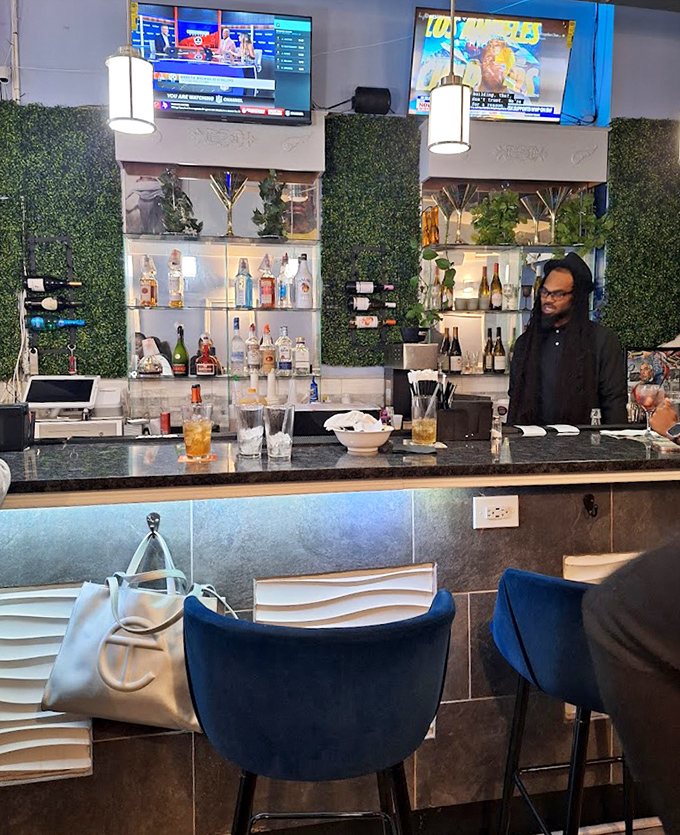 The bar area balances elegance and comfort with its green wall backdrop, gleaming bottles, and blue velvet chairs &ndash; cocktail hour never looked so inviting.