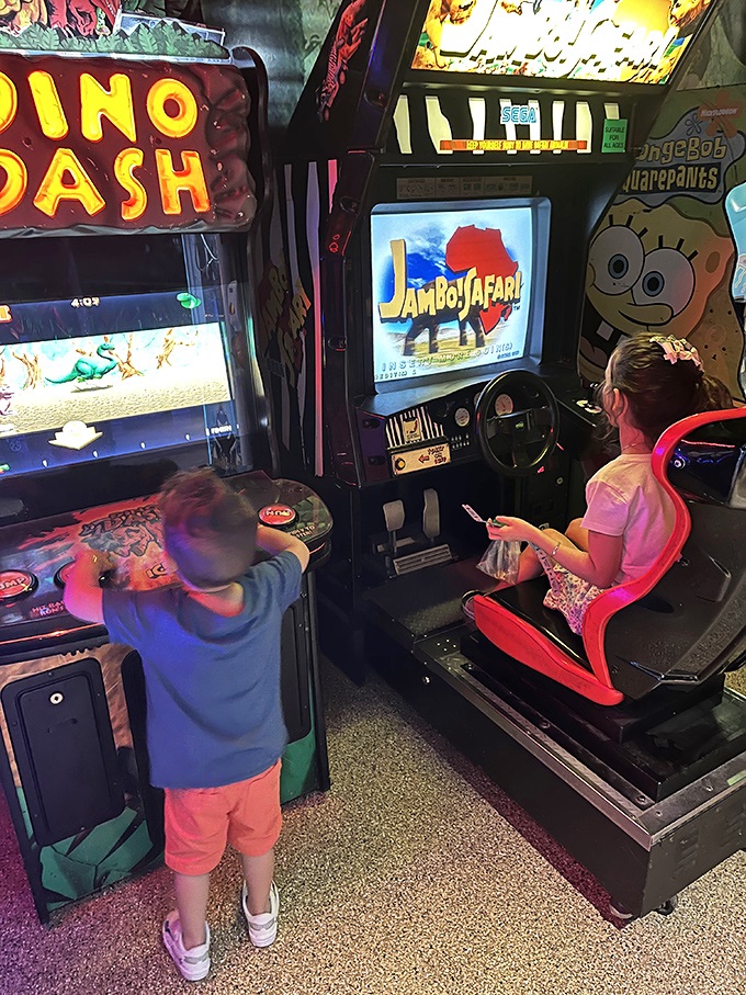 Digital dinosaurs await in the arcade area. Kids bounce between games with faces illuminated by screen glow as they navigate virtual prehistoric challenges.