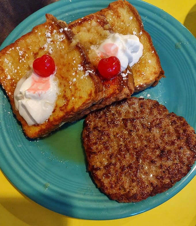 French toast topped with whipped cream and cherries alongside a perfectly seasoned sausage patty is breakfast goals personified.