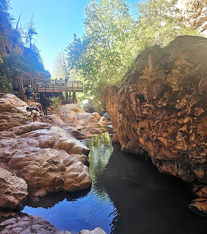 Sunlight dapples the creek below as visitors navigate rocky passages, each footstep a brief interaction with eons of geological history.