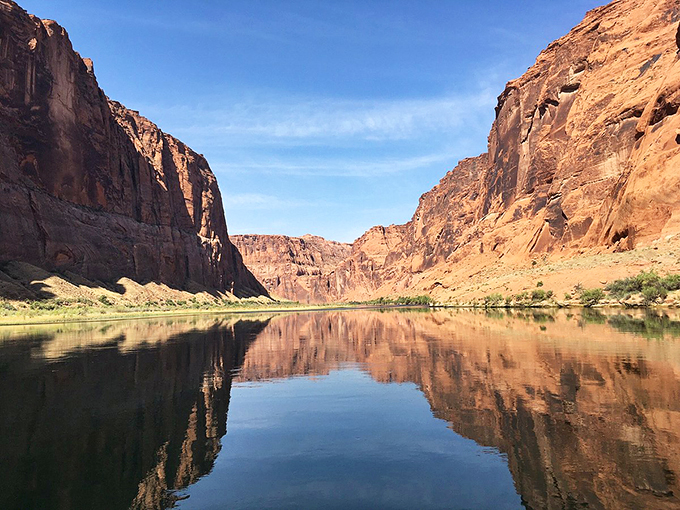 Red rock walls rise like ancient skyscrapers, reflecting their majesty in the calm Colorado River below.