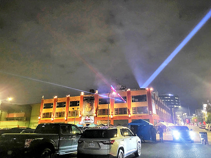 Parking Area: The illuminated building stands like a nightmare factory against the night sky, drawing in thrill-seekers like moths to flame.