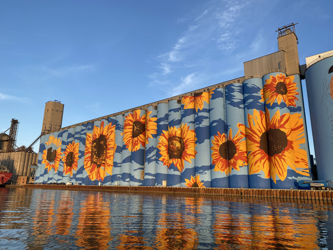 The Glass City River Wall catches golden hour light, its vibrant sunflowers reflecting in the Maumee River like nature's perfect mirror.