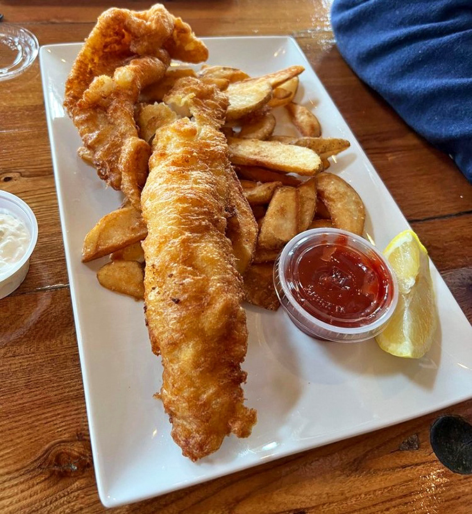 Golden-battered haddock stretches across the plate like a delicious treasure, accompanied by perfectly crisp fries and essential dipping sauces.