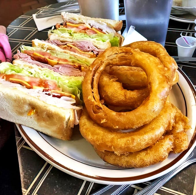 That club sandwich stands tall and proud, flanked by golden onion rings that shatter perfectly with each bite &ndash; a dynamic duo of deliciousness.