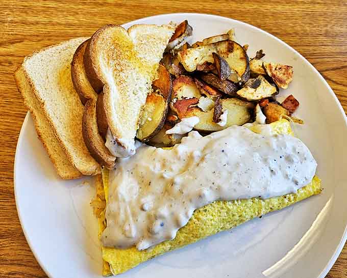 Omelet perfection that would make a French chef weep – fluffy eggs blanketed with creamy gravy, served with golden potatoes and toast.