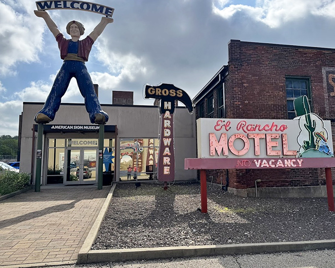 The American Sign Museum's exterior features a joyful figure holding a "Welcome" sign high above the entrance, while vintage motel and restaurant signs create a nostalgic roadside scene.