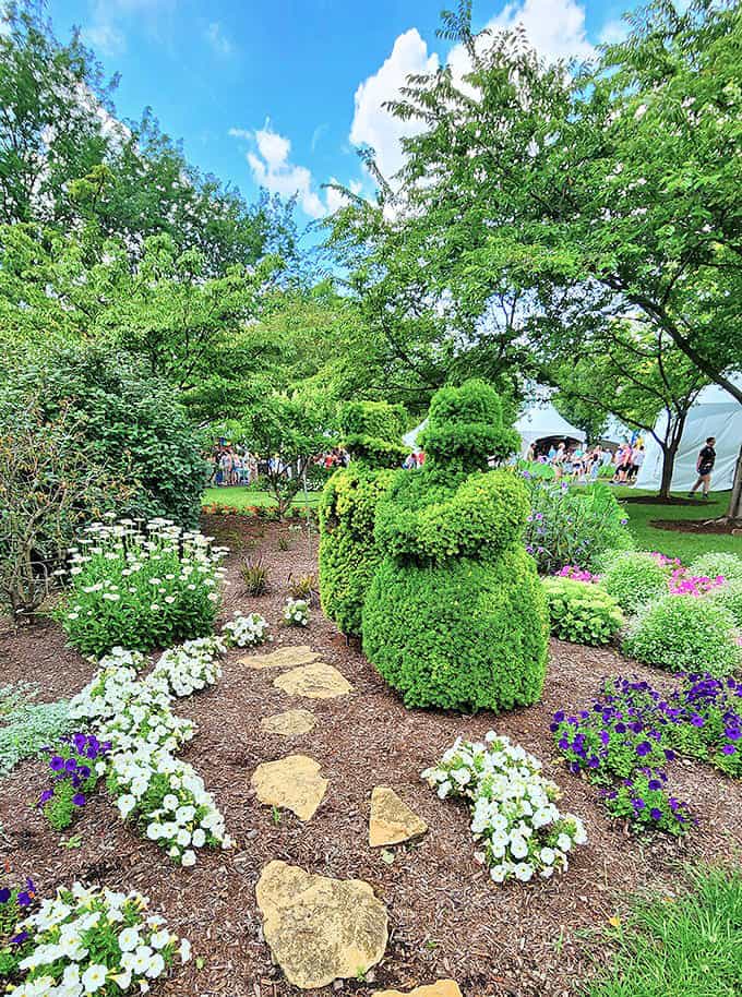 A whimsical topiary bear welcomes visitors along a path lined with cheerful white and purple blooms.