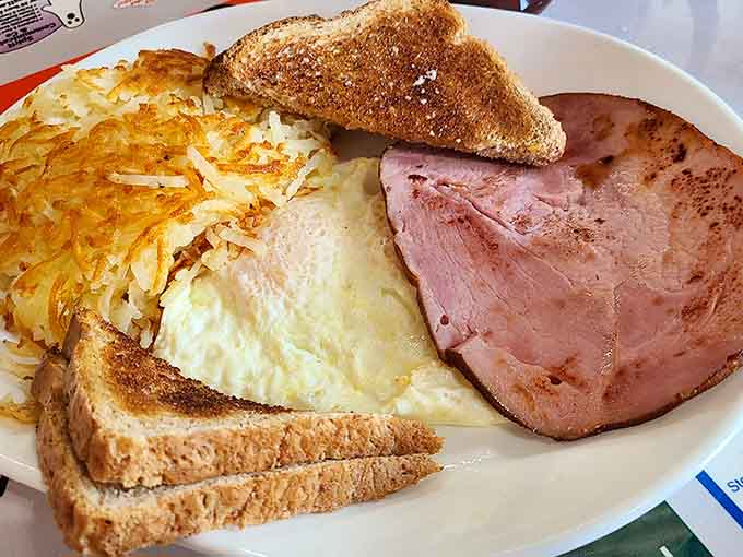 The classic breakfast plate arrives loaded with perfectly cooked eggs, crispy hash browns, thick ham, and toast that's actually toasted, not just warmed.