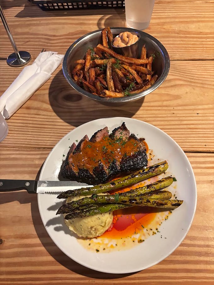A perfectly seared flat iron steak rests beside grilled asparagus and house fries &ndash; comfort food elevated without losing its soul.