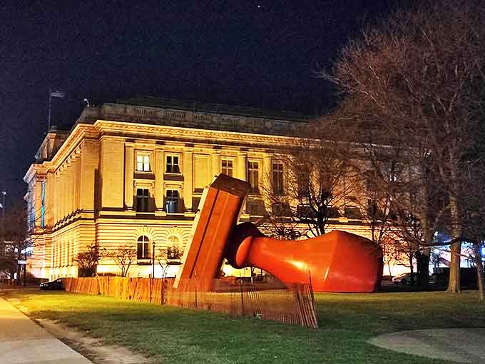 By night, the Free Stamp transforms into a dramatically lit urban monument, its red form glowing against the illuminated Cleveland cityscape.