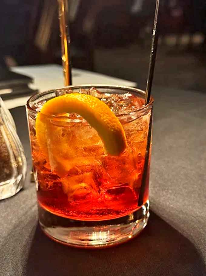 The Negroni glows amber in the dim light, its perfect balance of bitter Campari, sweet vermouth, and botanical gin garnished with an orange twist that adds aromatic magic.