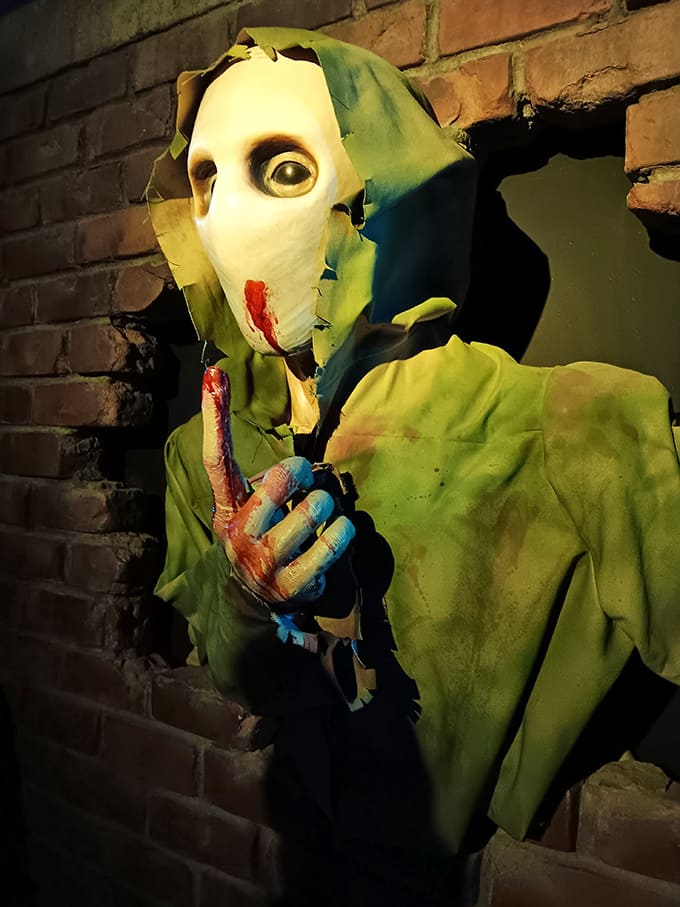 A chilling masked figure in tattered green clothing silently urges visitors to keep quiet, its stitched face and bloody hands promising unspeakable consequences.