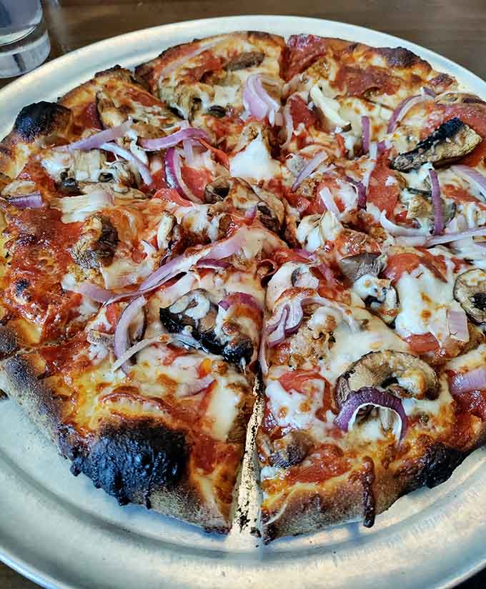 Wood-fired pizza with mushrooms, tomatoes, and red onions emerges from that copper-hooded oven with leopard-spotted crust perfection.