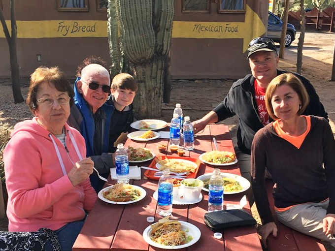 Happy diners gather around a picnic table laden with Indian Village specialties, proving good food brings people together in the Arizona sunshine.