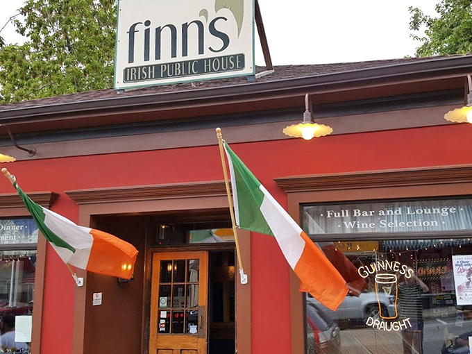 Two Irish flags flutter proudly outside this cozy red-painted pub, where warm golden lights promise good times and great food ahead.
