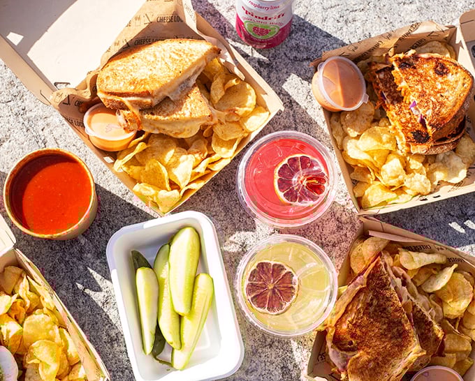 A picnic-perfect spread featuring golden-grilled sandwiches, fresh pickles, and drinks garnished with citrus slices. Photo credit: Cheese Louise Portland