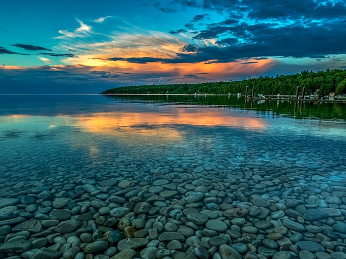 Crystal-clear water unveils a bed of smooth stones, reflecting a fiery sunset&mdash;nature&rsquo;s perfect blend of tranquility and vibrant color.