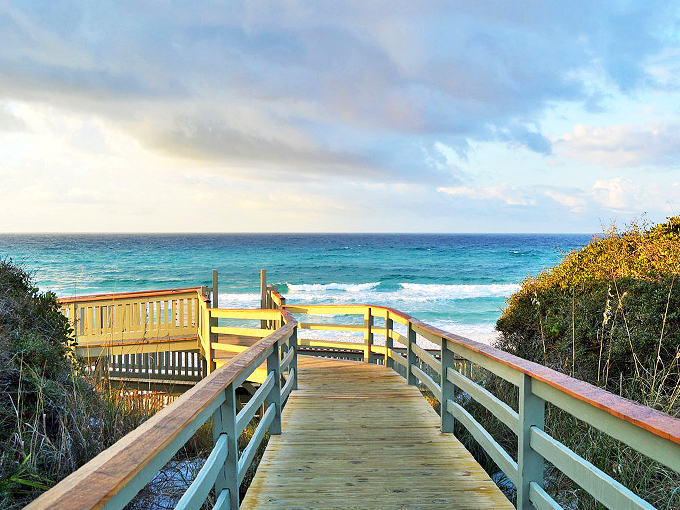 A wooden boardwalk leads to paradise, where turquoise waves meet powder-white sand under a pastel-painted sky.