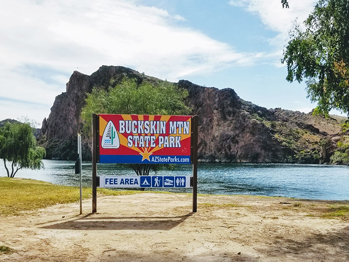 Buckskin Mountain State Park's entrance sign stands as a gateway to adventure, with the iconic red and blue signage framed by rugged mountain peaks and tranquil shoreline.