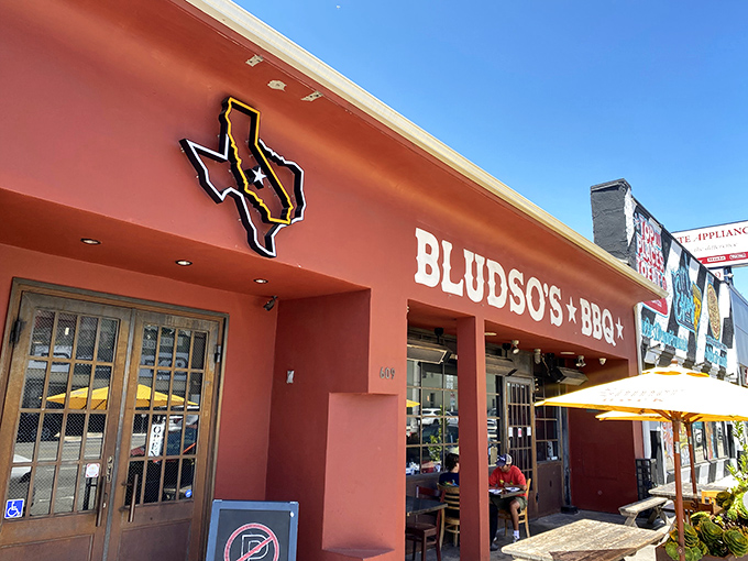 The bright red exterior of Bludso's BBQ stands like a beacon of hope for hungry Angelenos, promising smoky salvation under those cheerful yellow umbrellas.