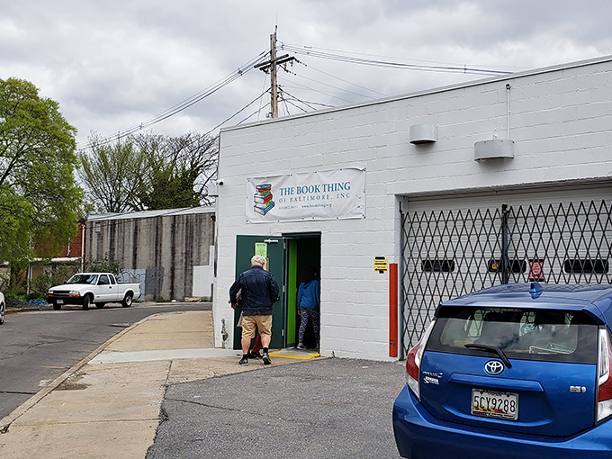 The unassuming exterior of literary heaven – a white cinder block building with a simple banner that promises bookish treasures beyond the green door.