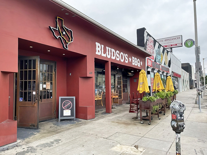 The bright red exterior of Bludso's BBQ stands like a beacon of hope for hungry Angelenos, promising smoky salvation under those cheerful yellow umbrellas.