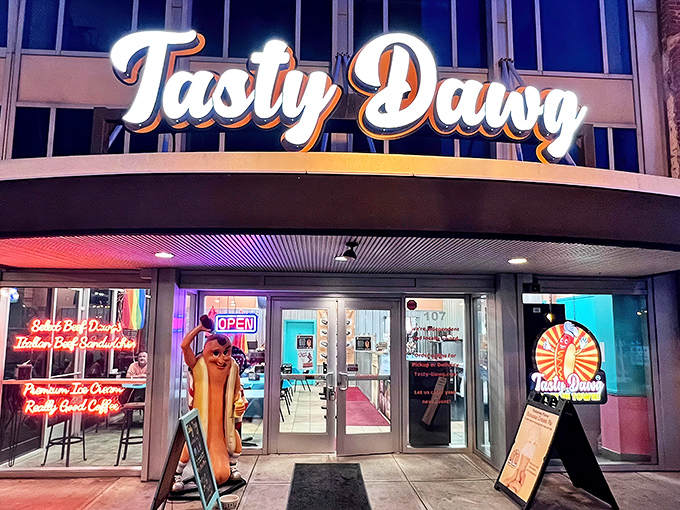 The glowing beacon of Tasty Dawg welcomes hungry patrons with its vibrant signage and life-sized hot dog mascot standing guard, promising delicious adventures within.