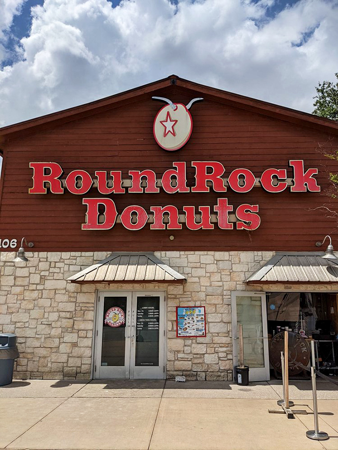 The iconic wooden and limestone facade of Round Rock Donuts stands proudly under Texas skies, beckoning donut pilgrims with its famous red signage.
