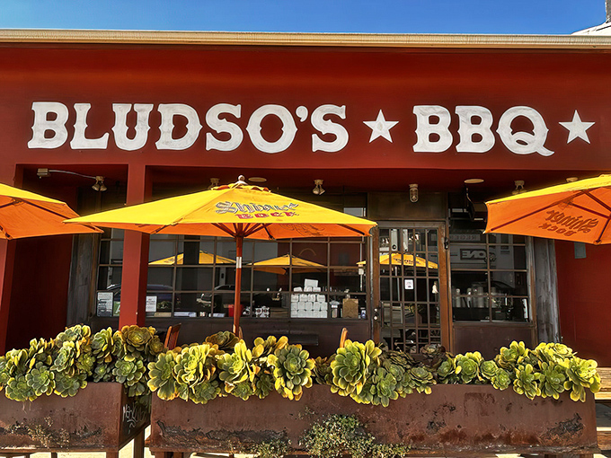 The bright red exterior of Bludso's BBQ stands like a beacon of hope for hungry Angelenos, promising smoky salvation under those cheerful yellow umbrellas.