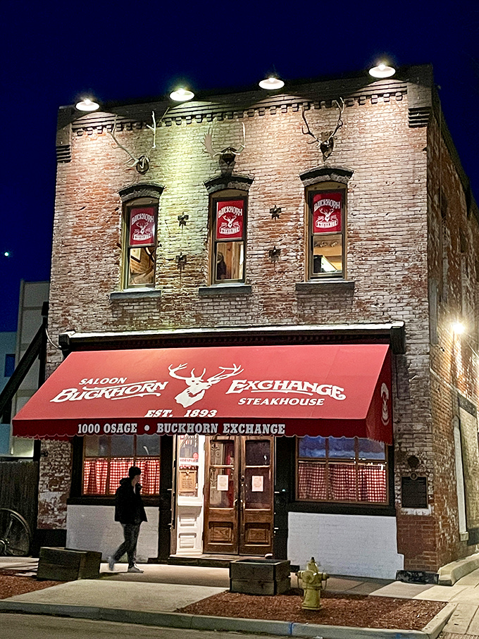 The brick facade of Buckhorn Exchange glows under night lights, its iconic red awning beckoning hungry travelers like a carnivorous lighthouse in urban Denver.