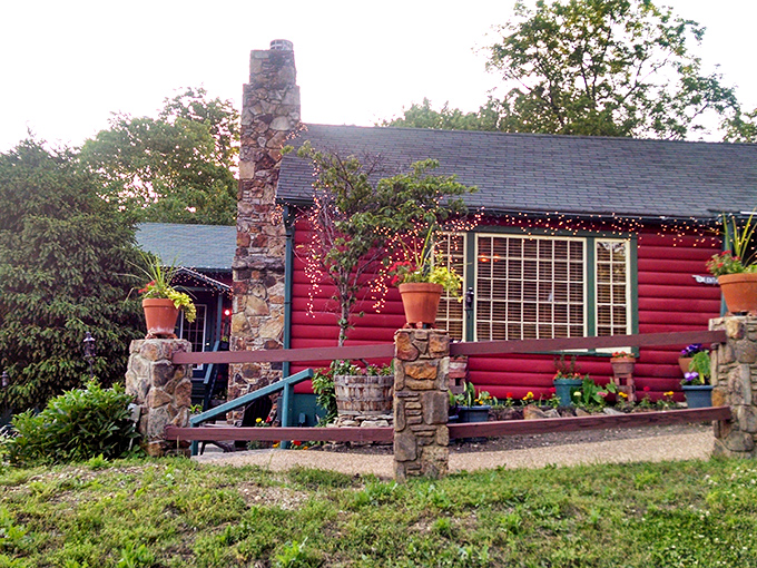 The crimson log cabin emerges from lush Ozark greenery like a storybook setting—complete with stone chimney and verdant landscaping that whispers "come on in."