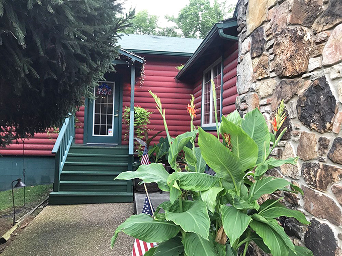 The crimson log cabin emerges from lush Ozark greenery like a storybook setting&mdash;complete with stone chimney and verdant landscaping that whispers "come on in."
