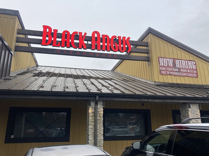 The glowing red signage of Black Angus beckons hungry travelers like a carnivorous lighthouse guiding ships to beefy salvation.