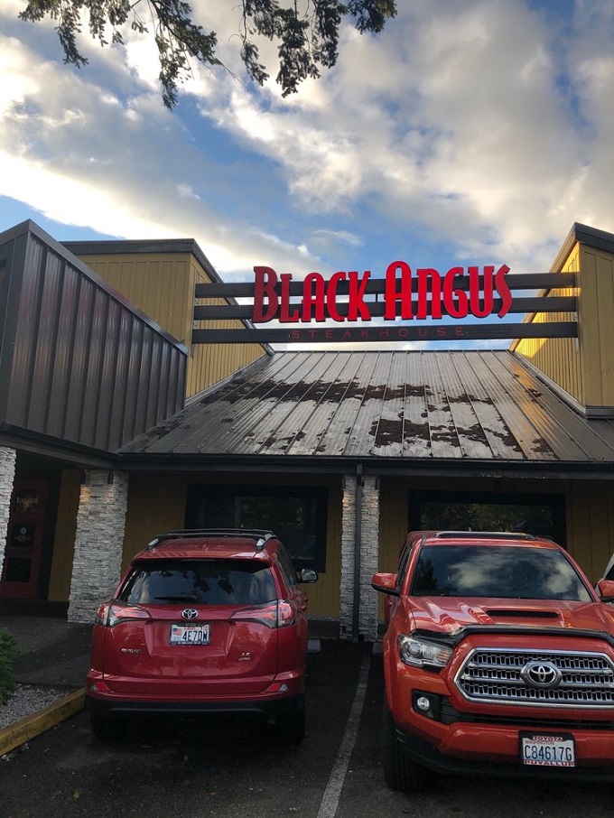 The glowing red signage of Black Angus beckons hungry travelers like a carnivorous lighthouse guiding ships to beefy salvation.