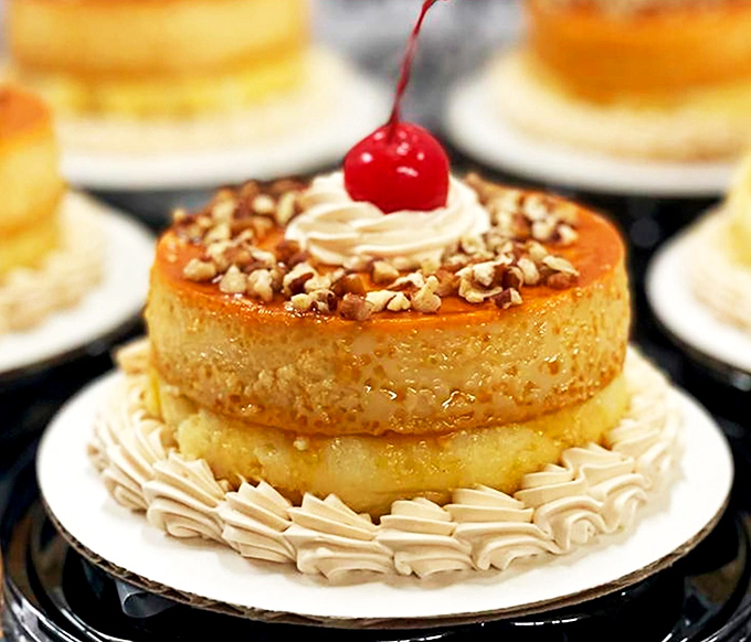 The flan cheesecake sits like royalty on its throne of whipped cream, the caramel crown and cherry scepter signaling its dessert sovereignty.