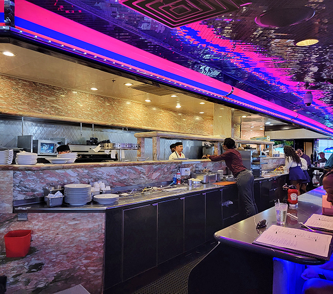 The kitchen buzzes with the choreographed precision of a Cirque du Soleil show, all under the glow of that signature neon pink lighting.