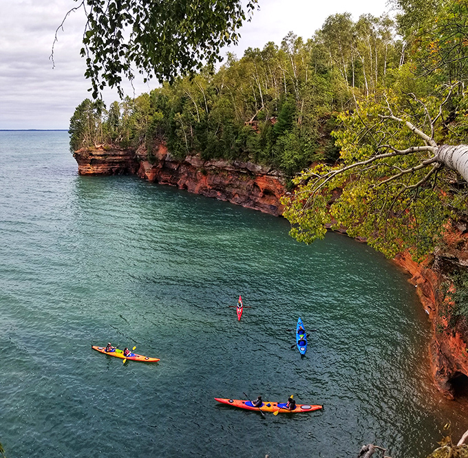 Kayakers glide beneath towering red cliffs like explorers in a forgotten world, dwarfed by nature's majesty in the Apostle Islands.
